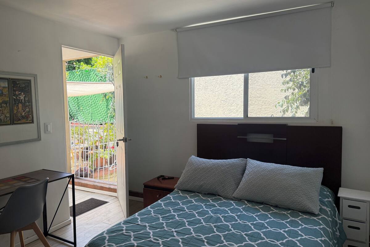 Quarto com cama, escrivaninha, cadeira, janela e porta que dá para área externa.