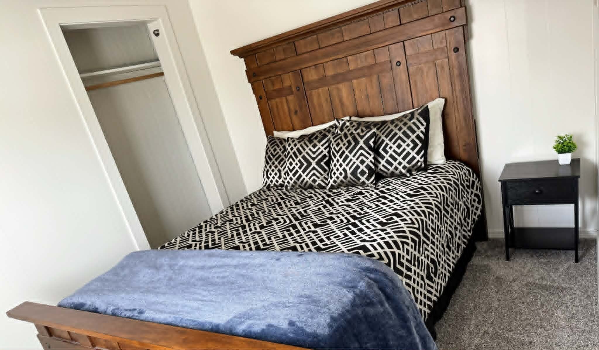 Quarto com cabeceira de madeira, roupa de cama estampada, mesa de cabeceira e uma pequena planta.