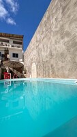 Uma piscina com água azul cristalina, um edifício com parede texturizada e um guarda-sol vermelho.