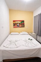 Uma cama de solteiro com lençóis brancos, um quadro com uma cena de pôr do sol na parede e uma cortina cinza na janela.