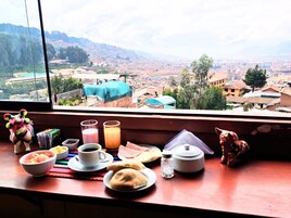 Mesa de café da manhã com comida e bebidas, janela com vista para a cidade.