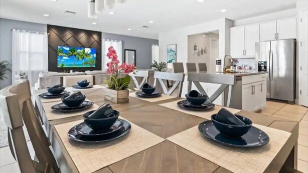Uma sala de jantar moderna com uma mesa grande posta para seis pessoas, uma cozinha com armários brancos e uma TV de tela plana na parede.