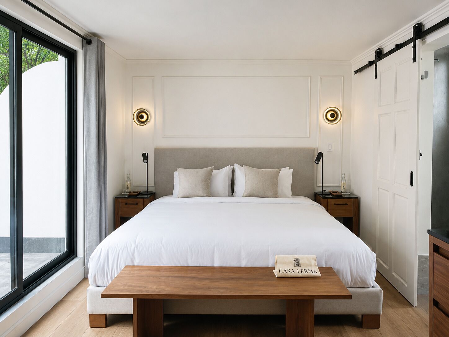 Quarto moderno com uma cama grande, mesas de cabeceira, um criado-mudo de madeira, um banco e uma porta deslizante.
