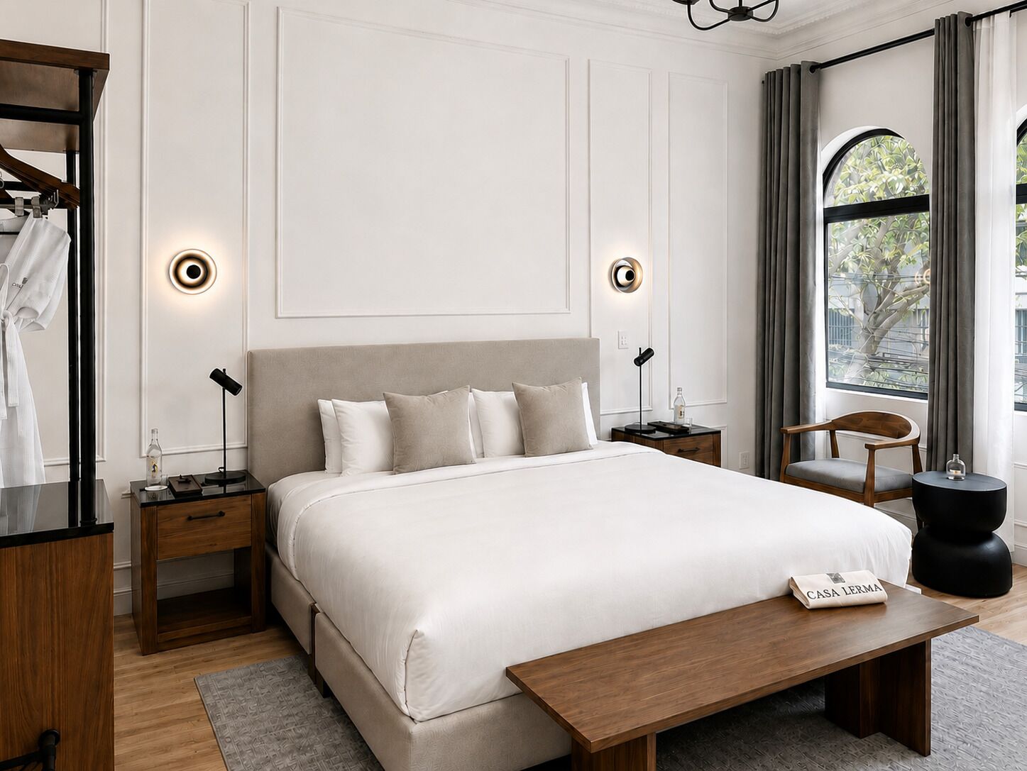Quarto de hotel com uma cama grande, mesas de cabeceira, um criado-mudo, uma cadeira, uma pequena mesa e uma janela com cortinas.