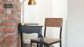 Quarto de canto com parede de tijolos, uma escrivaninha com abajur, uma cadeira e um tapete estampado.