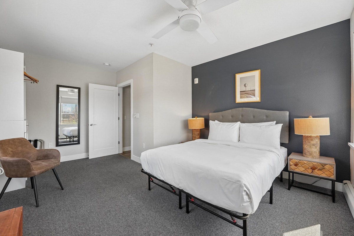 Quarto de hotel com cama, duas luminárias de cabeceira, uma cadeira, guarda-roupa e ventilador de teto.