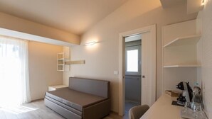 Quarto moderno com uma cama, uma escrivaninha e uma cadeira. Há uma porta que dá para outro cômodo.