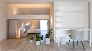 Quarto moderno e bem iluminado, com uma cama, mesa de cabeceira e escrivaninha com cadeira. Há plantas em vasos no chão.