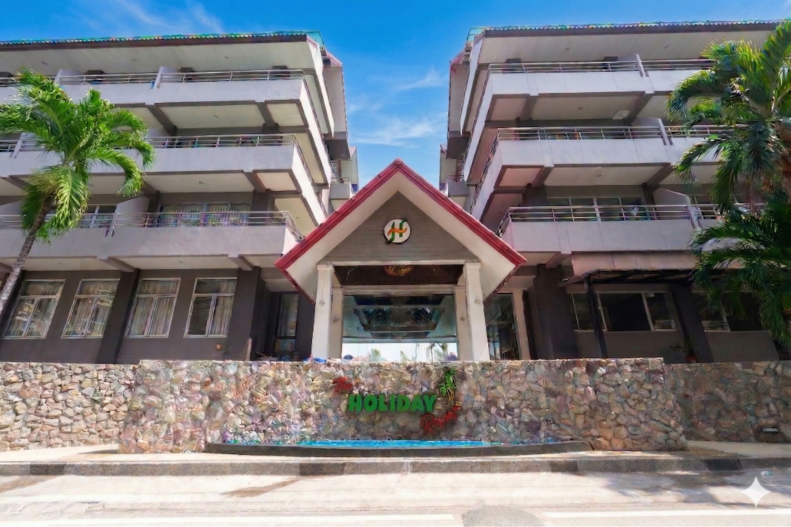 Um edifício de vários andares com varandas, um muro de pedra com a placa 'HOLIDAY' e palmeiras.
