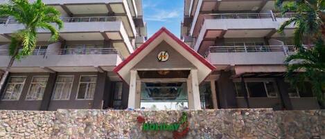 Um edifício de vários andares com varandas, um muro de pedra com a placa 'HOLIDAY' e palmeiras.