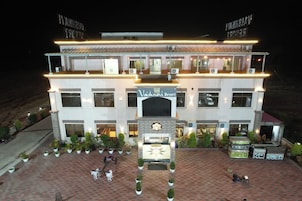 Um edifício de dois andares com uma placa indicando 'Vaishnavi Resort'. Há vários aparelhos de ar condicionado, plantas em vasos e uma pequena área de estar ao ar livre.