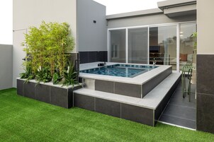 Uma área de piscina moderna ao ar livre, com hidromassagem, rodeada por azulejos escuros e grama sintética.