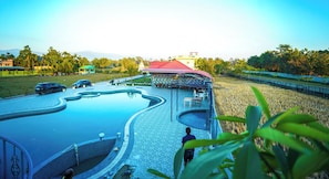 Piscina com um pavilhão de telhado vermelho, área gramada e um edifício ao longe.