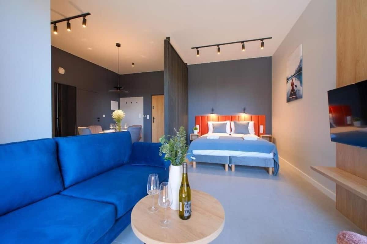 Quarto de hotel moderno com um sofá azul, uma cama com cabeceira laranja, uma mesa de centro de madeira com uma garrafa e copos, e uma TV na parede.