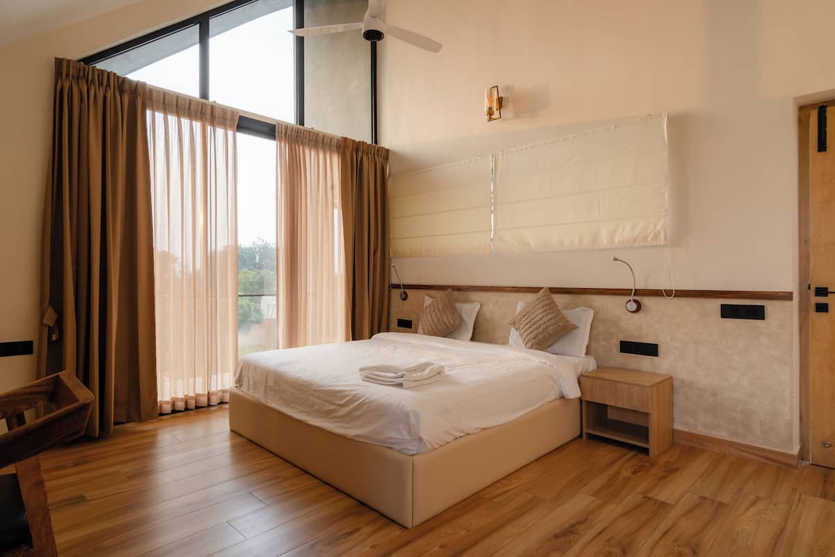 Quarto de hotel moderno com uma cama grande, piso de madeira, ventoinha no teto e vista pela janela.