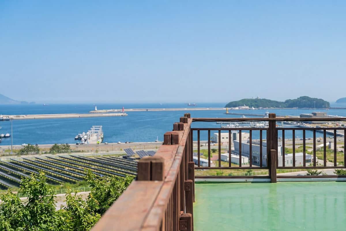 Vista costeira a partir de uma varanda, com o mar, um porto de recreio e colinas ao longe.