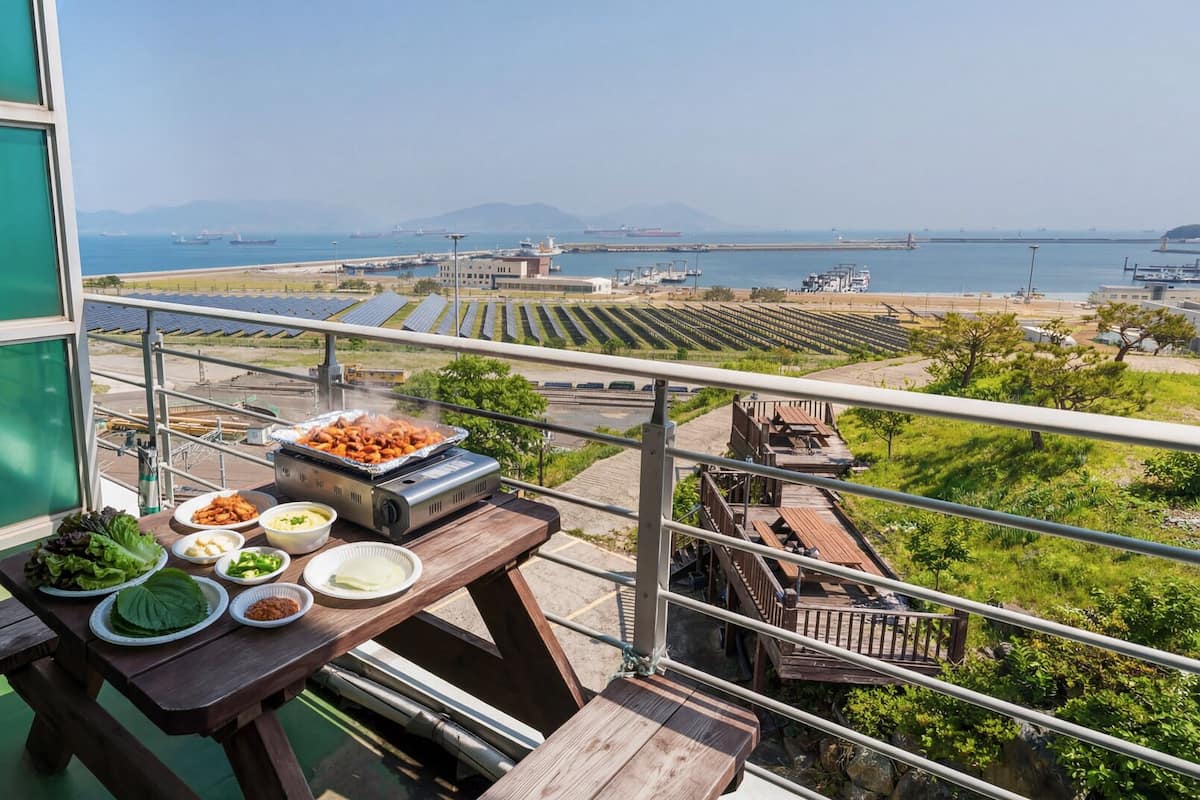 Uma varanda com vista para um porto, uma churrasqueira com comida e uma mesa com pratos.