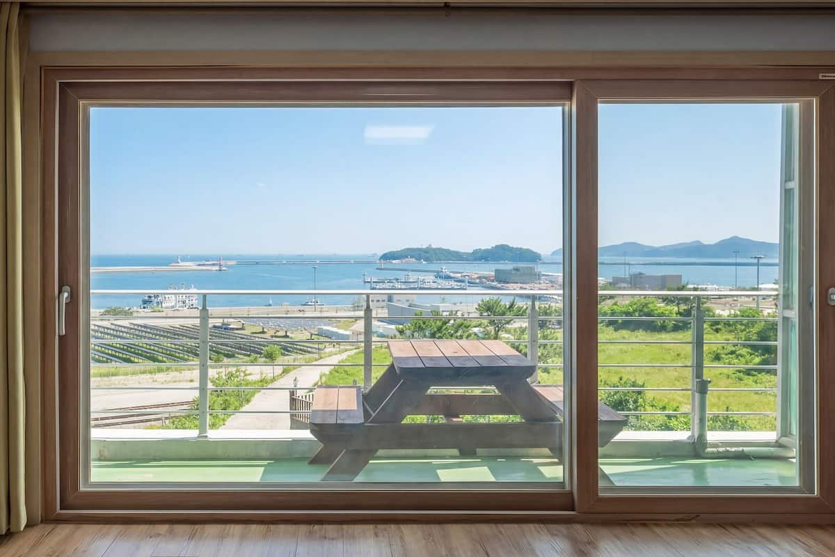 Quarto com porta de vidro deslizante que dá para uma varanda com vista para o mar, mesa de piquenique e guarda-corpo de metal.
