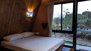 Quarto com cabeceira de madeira, cama com roupa de cama branca, mesa de cabeceira e janela com vista para pedras e vegetação.