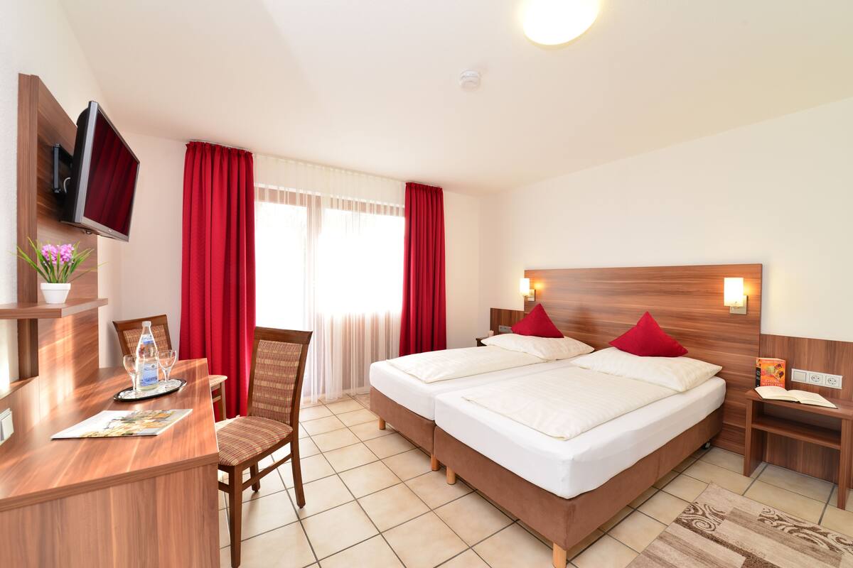 Quarto de hotel com cama, mesas de cabeceira, escrivaninha com cadeira, televisão e janela com cortinas.
