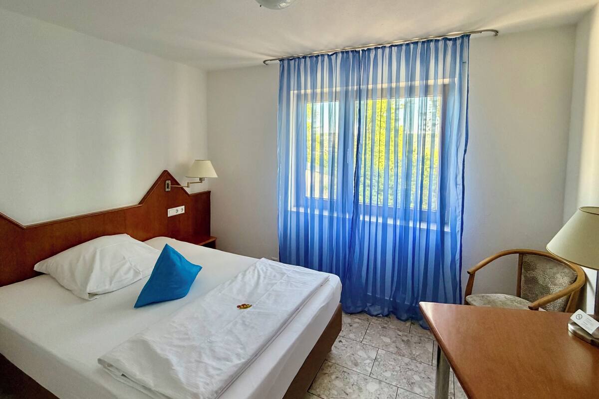 Quarto de hotel com cama, mesa de cabeceira de madeira, escrivaninha, cadeira e janela com cortinas azuis.