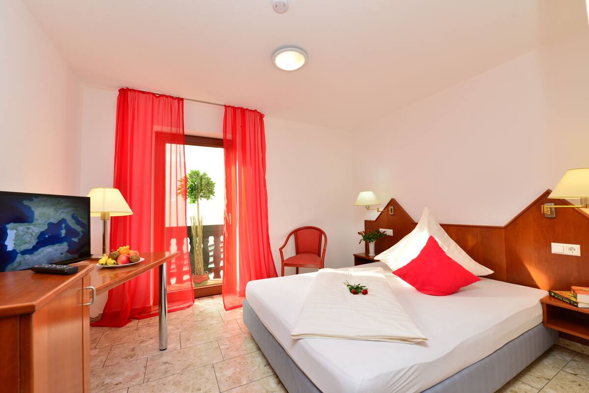 Quarto de hotel com cama, escrivaninha com televisão, cadeira, mesa de cabeceira e janela com cortinas vermelhas.