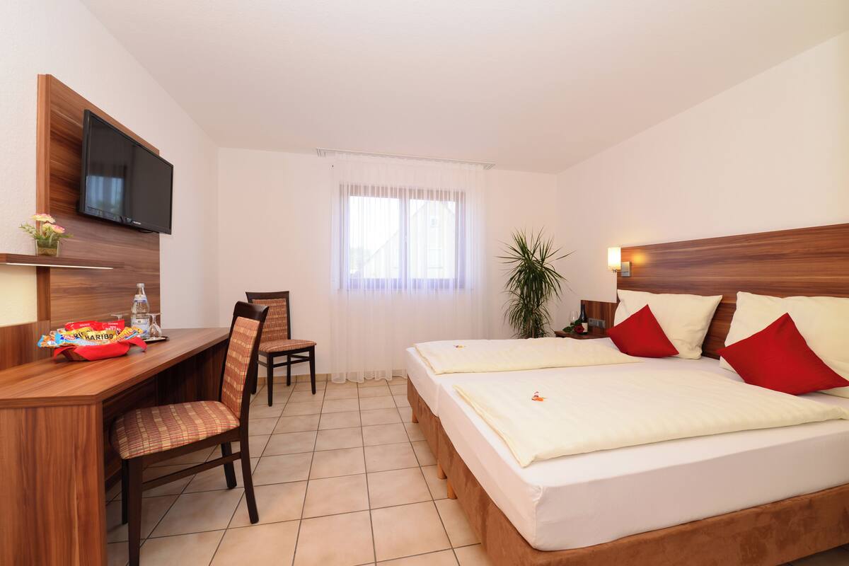 Quarto de hotel com uma cama, uma escrivaninha com cadeira, televisão, janela com cortinas e uma planta.