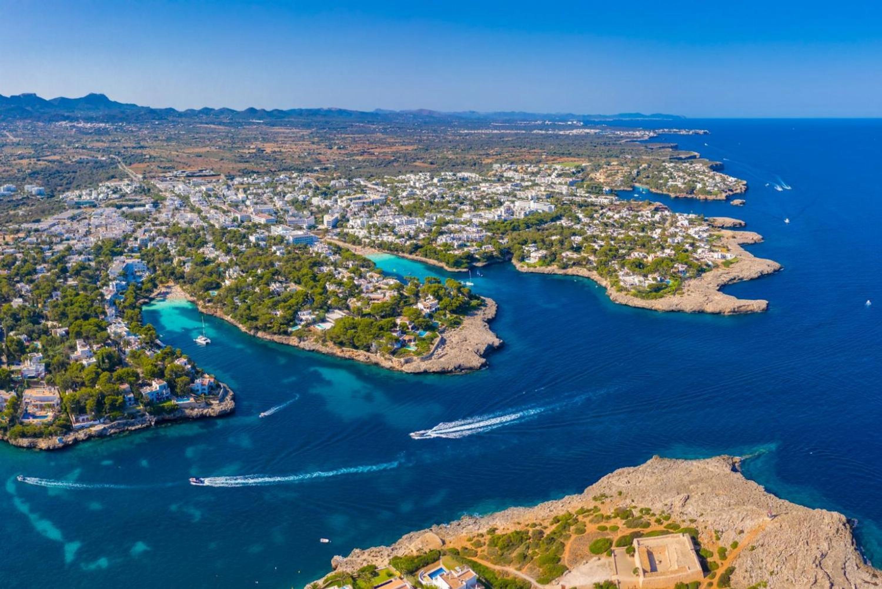 Vista aérea de uma cidade costeira com edifícios ao longo da orla, barcos na água e céu claro.