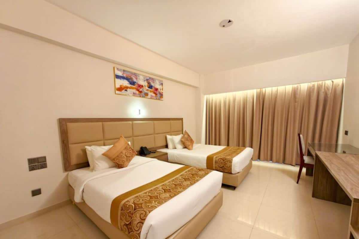 Quarto de hotel com duas camas, uma mesa, uma cadeira, um quadro na parede e uma janela com cortinas.