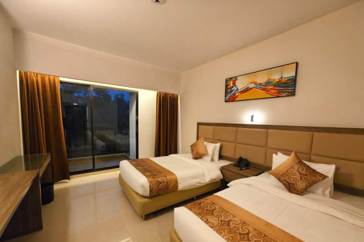 Quarto de hotel com duas camas, uma escrivaninha, uma cadeira, uma janela grande com cortinas e um quadro na parede.