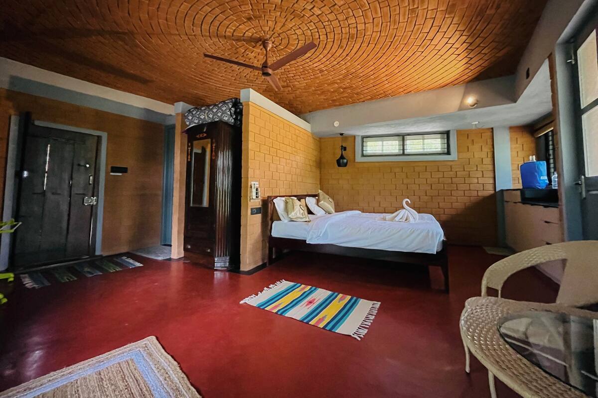 Um quarto com teto de tijolos, cama com lençóis brancos, porta de madeira e uma pequena mesa com uma cadeira.