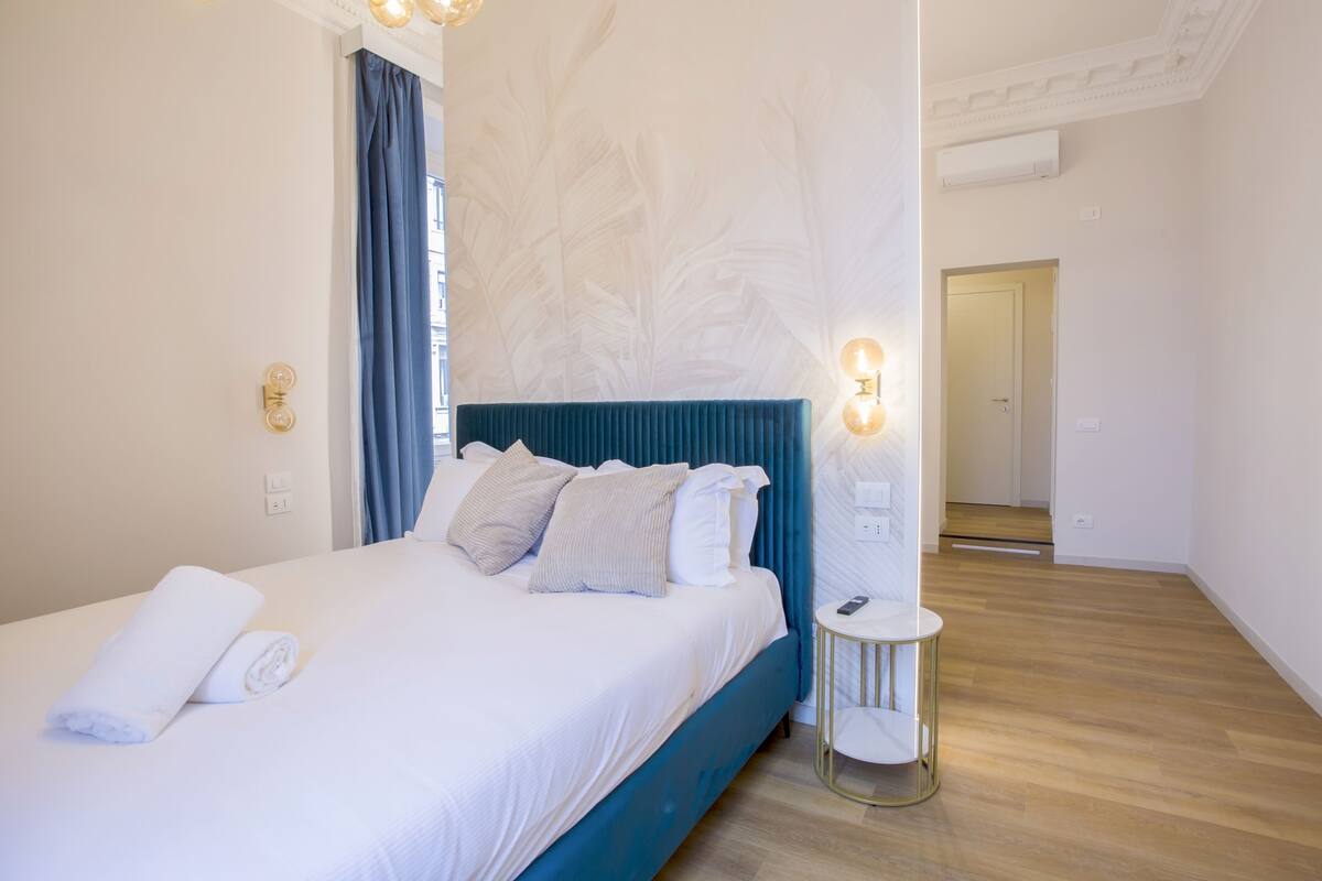 Quarto de hotel com cama, mesa lateral redonda, luminária na parede e porta que dá para outro cômodo.