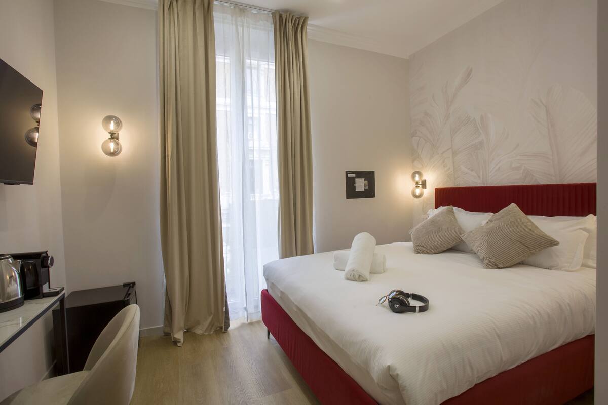 Quarto de hotel com cama, escrivaninha, cadeira, televisão e janela com cortinas.