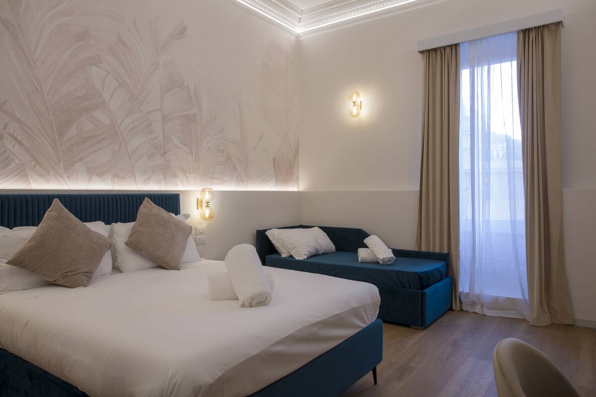 Quarto de hotel com cama grande, sofá, luminária na parede e janela com cortinas.