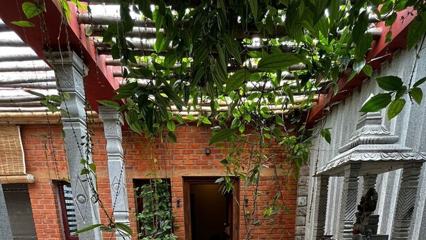 Uma trilha de pedra leva a uma porta, cercada por vegetação e elementos arquitetônicos tradicionais.
