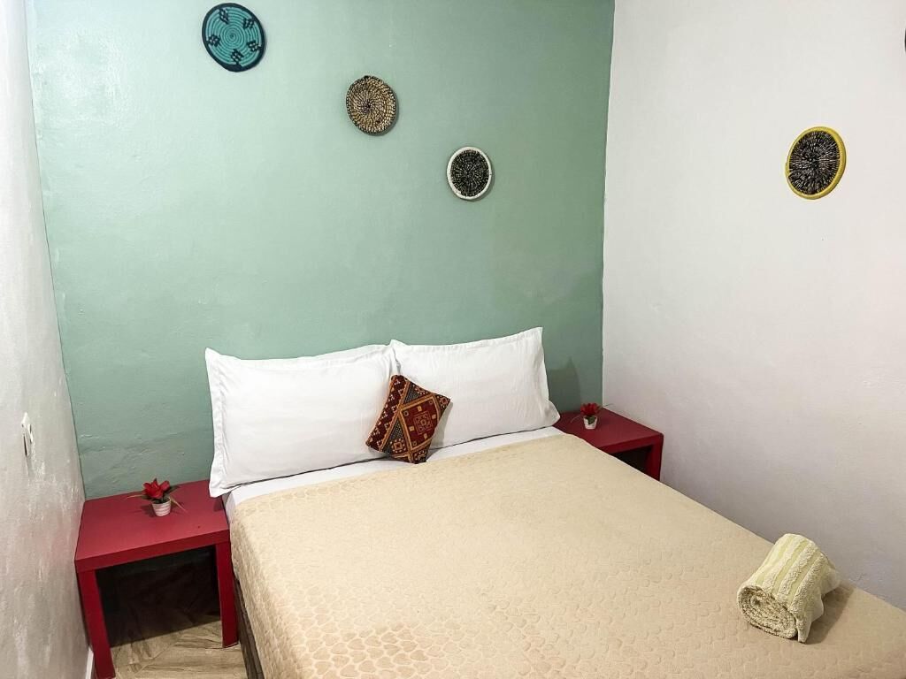 Quarto com cama, duas mesas de cabeceira, uma mesinha lateral vermelha e pratos decorativos na parede.