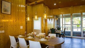 Uma sala de jantar com painéis de madeira, uma mesa grande posta para refeição, cadeiras brancas e luminárias pendentes.