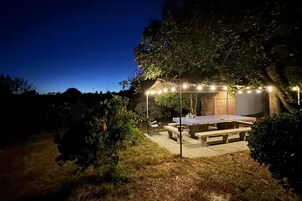 Um pátio externo com mesa e bancos, cercado por árvores e uma estrutura de tijolos.