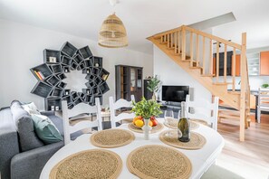 Uma sala de estar moderna com uma mesa de jantar, uma obra de arte geométrica na parede, uma escada e uma televisão ao fundo.