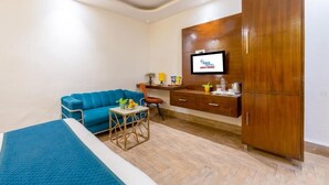 Quarto de hotel com uma cama, um sofá azul, uma mesinha com uma planta, uma TV fixada em um armário de madeira e um guarda-roupa de madeira.