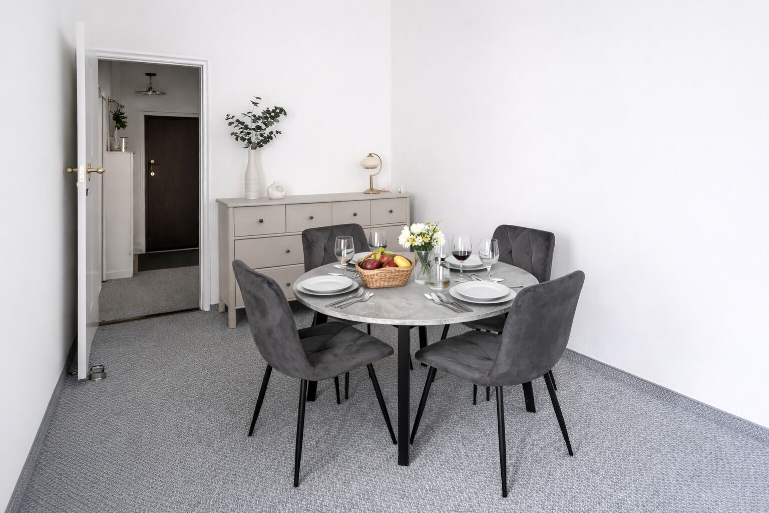 Uma sala de jantar com uma mesa redonda, quatro cadeiras cinzas, um vaso branco com flores, uma cesta de frutas e copos de vinho.