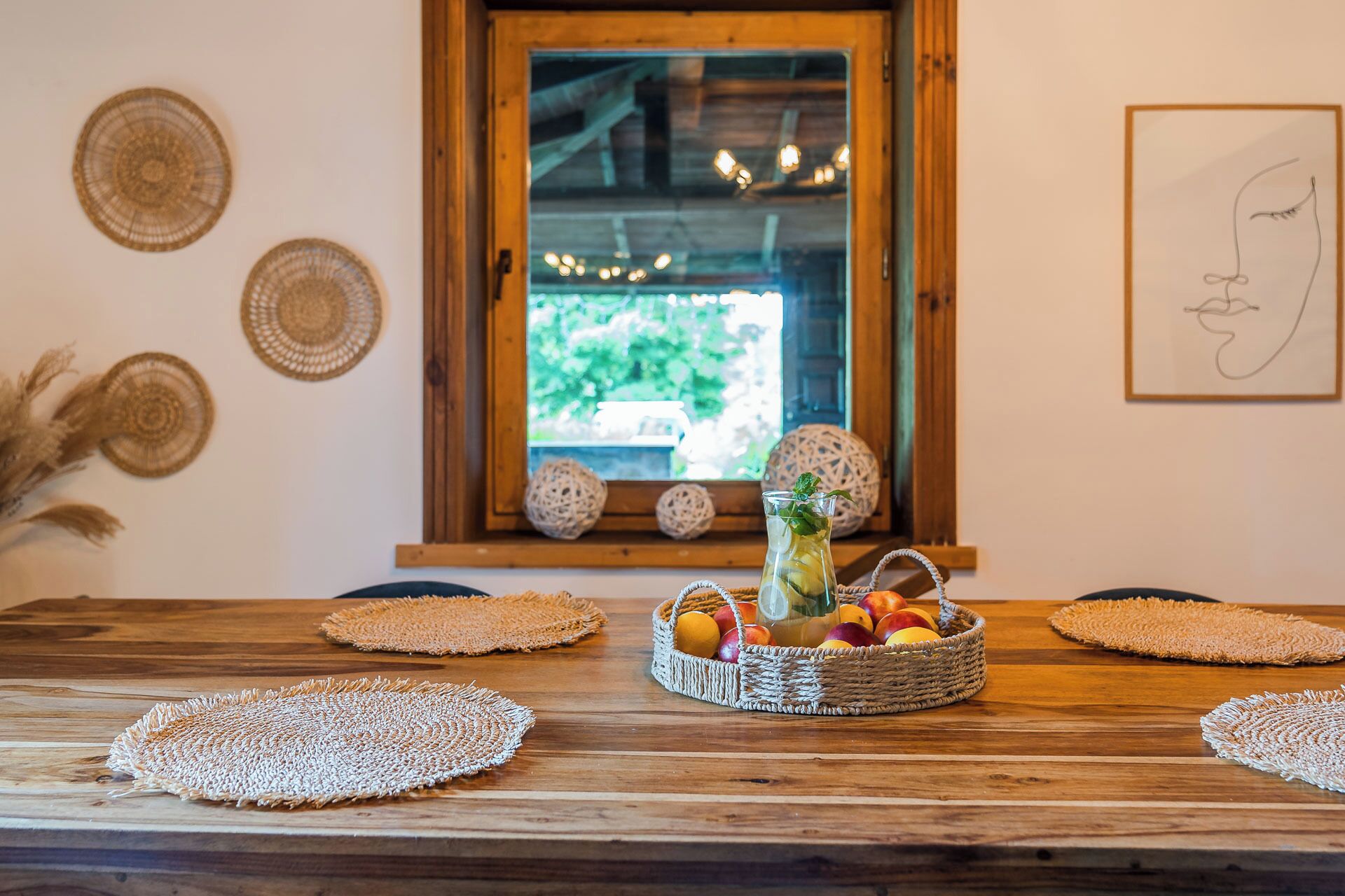 Uma mesa de madeira com jogos americanos trançados, uma cesta de frutas e um vaso com uma bebida.