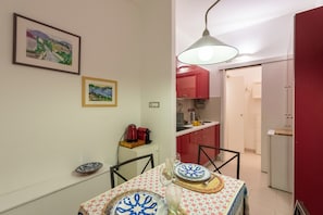 Uma área de refeições com uma mesa posta para dois, um armário vermelho ao fundo e uma cozinha visível através de uma porta aberta.