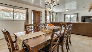 Uma sala de jantar com mesa de madeira, cadeiras, um lustre e uma televisão ao fundo.