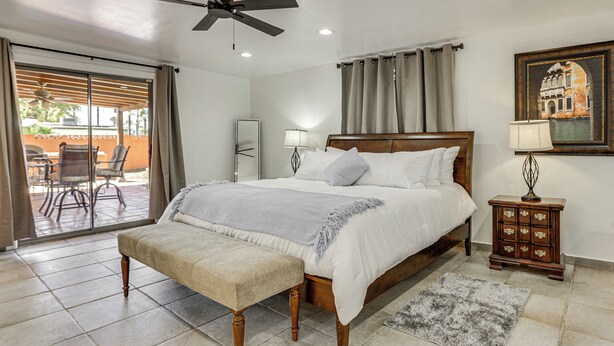 Quarto com cama, um banco, ventilador de teto, mesa de cabeceira e vista para área externa por uma porta deslizante.