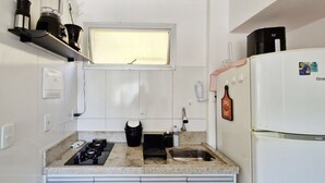Cozinha compacta com fogão a gás, pia, geladeira e janela com persianas.