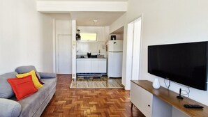 Sala de estar com sofá, televisão e cozinha ao fundo.