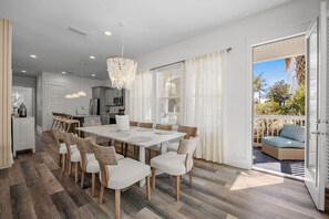 Cozinha moderna com armários brancos, área de refeições com mesa e cadeiras brancas e vista para uma varanda com mobiliário de exterior.