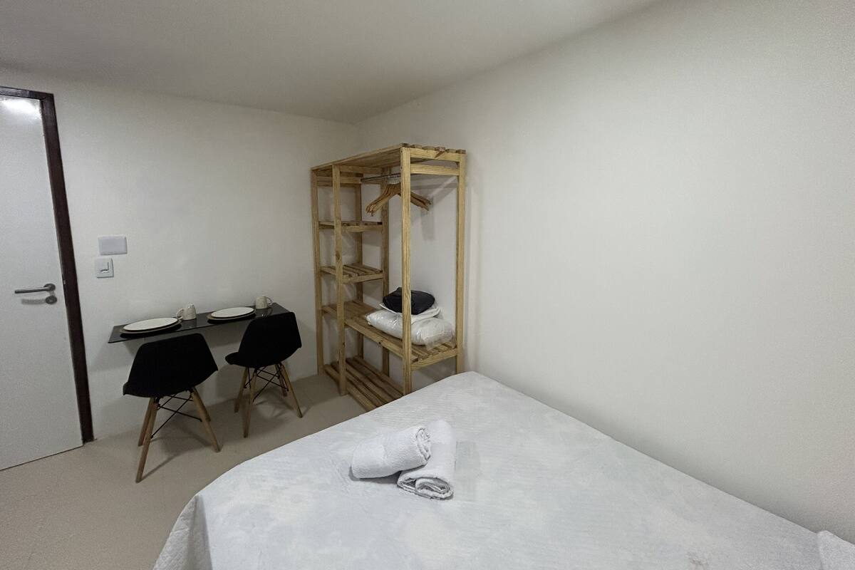 Quarto pequeno com uma cama de solteiro, beliche de madeira, escrivaninha e uma cadeira, além de uma porta.