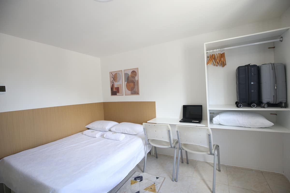 Quarto de hotel com cama, escrivaninha, cadeira, laptop e guarda-roupa com roupas penduradas.
